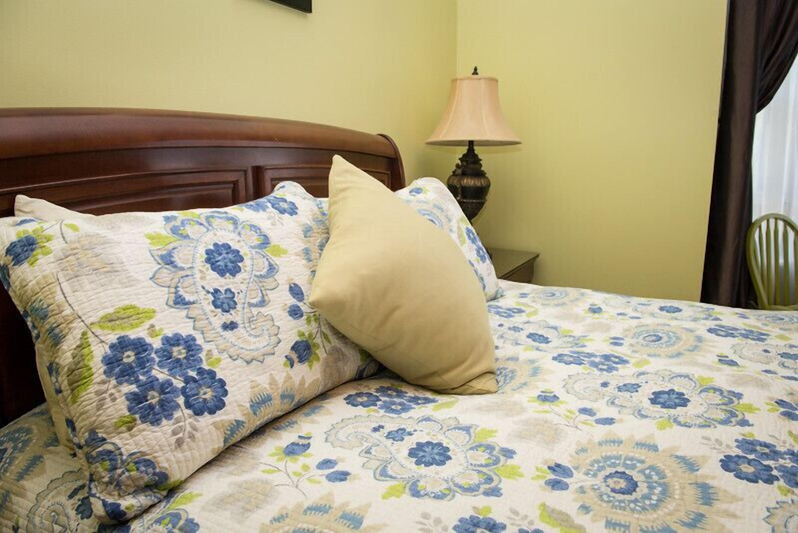 Cama com colcha florida e duas almofadas, cabeceira de madeira, luminária de cabeceira e uma cadeira em quarto com paredes amarelas.