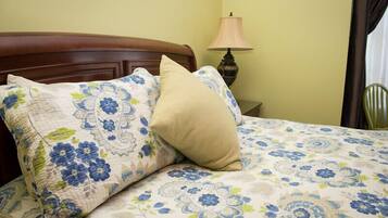 Cama com colcha florida e duas almofadas, cabeceira de madeira, luminária de cabeceira e uma cadeira em quarto com paredes amarelas.