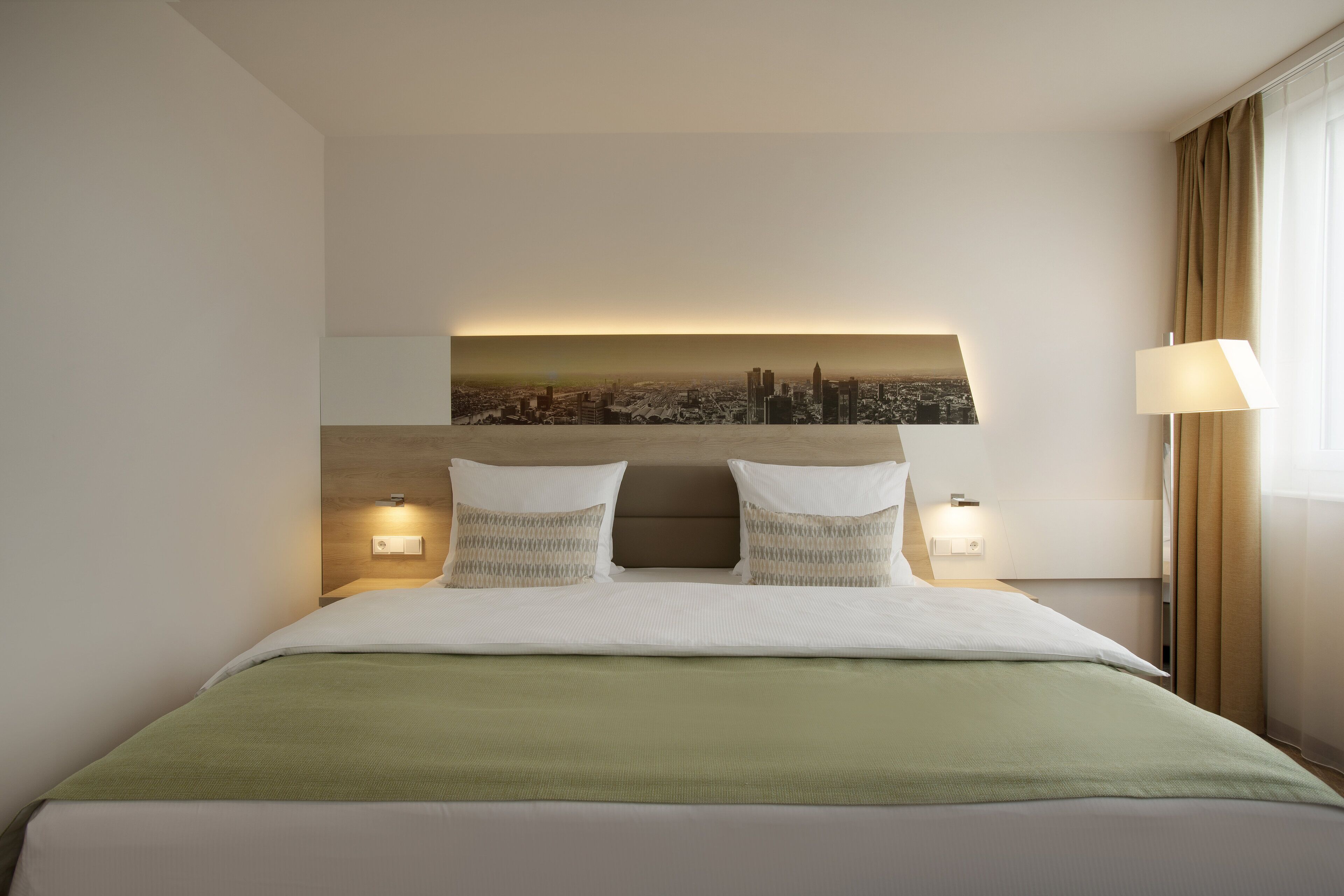 Quarto de hotel com uma cama, um quadro com uma vista urbana na parede e um abajur lateral.