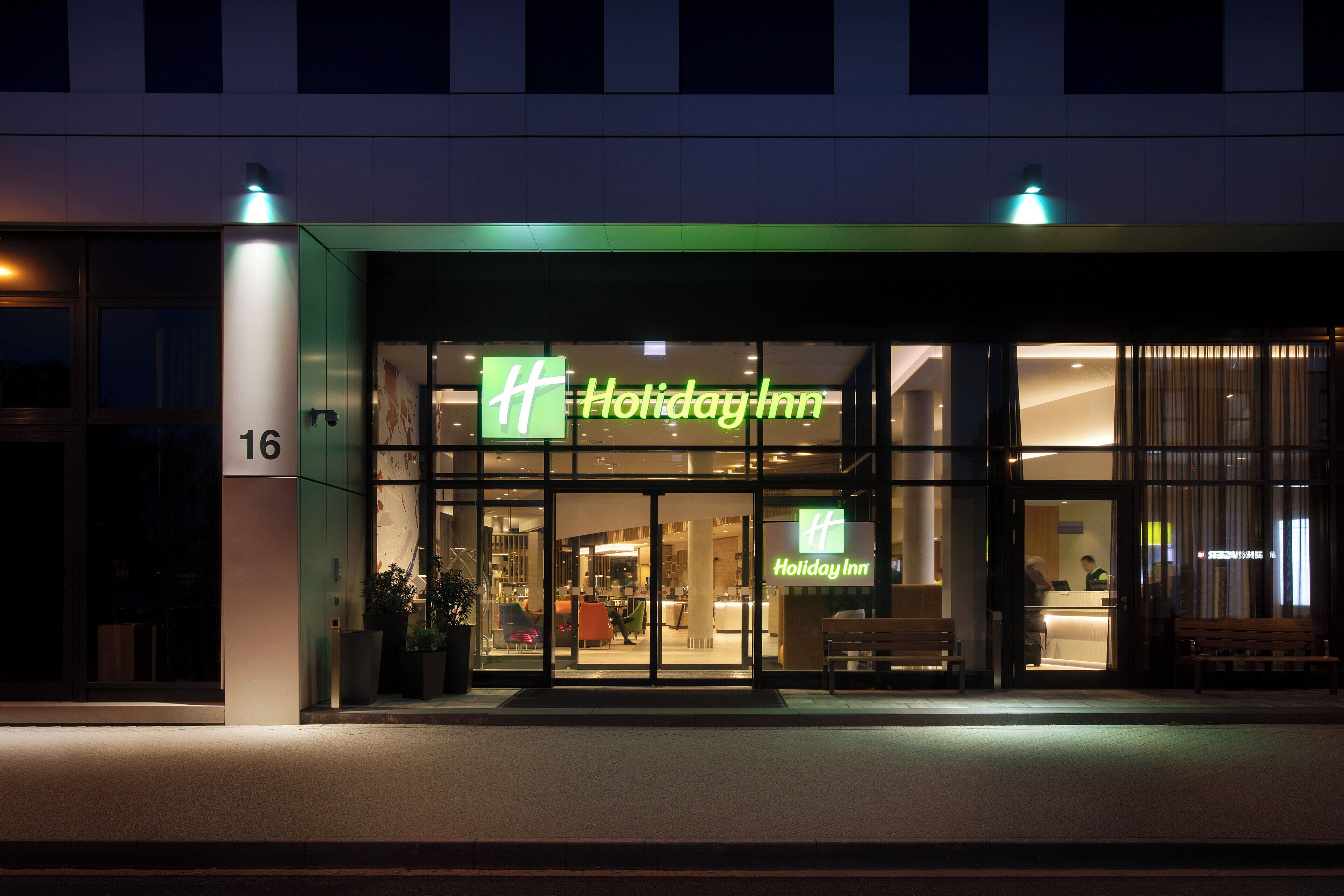 Entrada de um hotel Holiday Inn com fachada de vidro, balcão de recepção e área de estar ao ar livre.