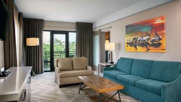 Quarto de hotel com sofá, mesa de centro, televisão, pintura e vista para o verde pela janela.