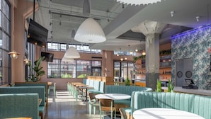 Um café moderno com bancos verdes e mesas redondas, janelas amplas e diversas plantas.