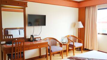 Quarto de hotel com escrivaninha de madeira, duas cadeiras, televisão, abajur e uma cama.