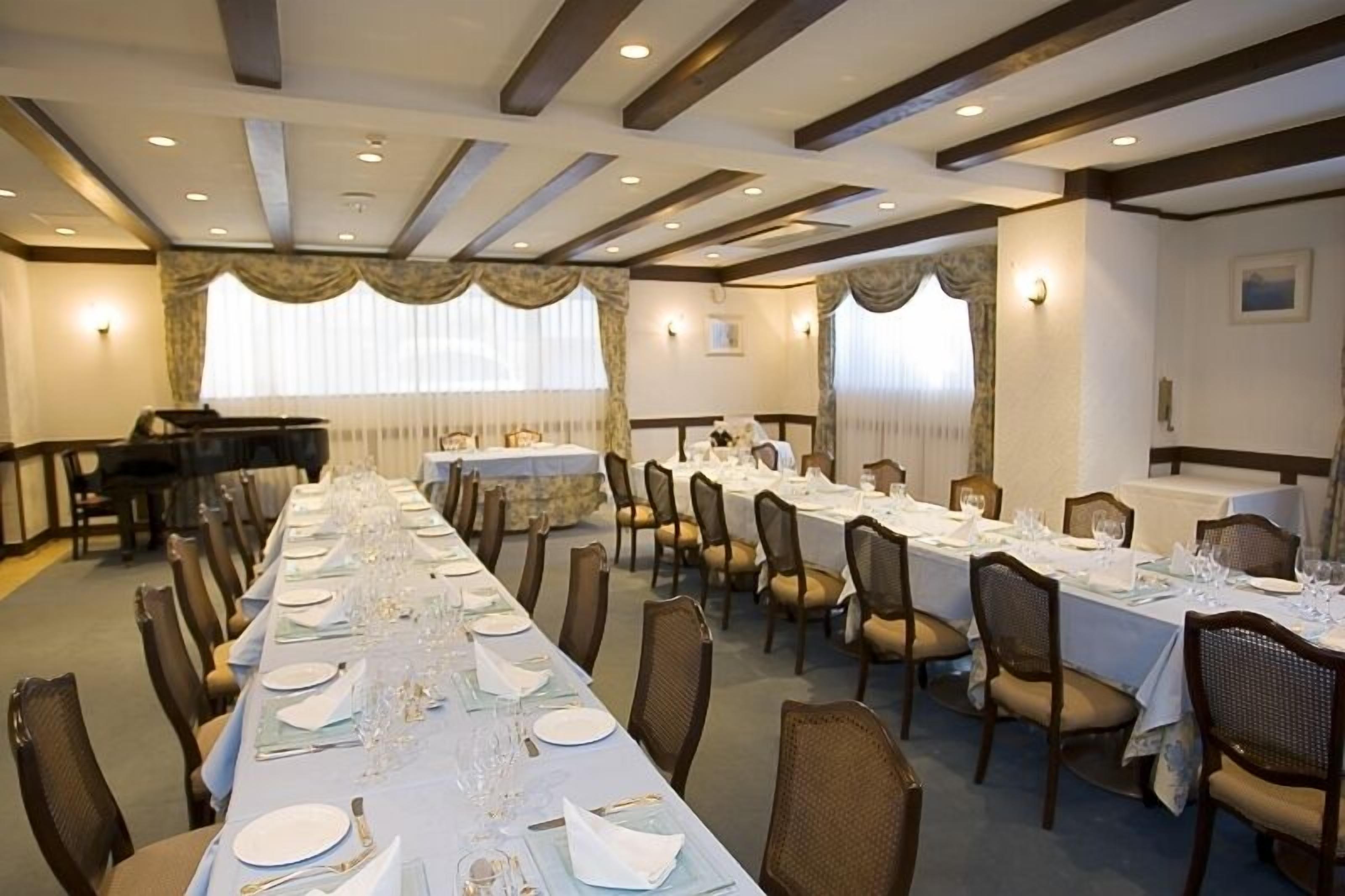 Uma sala de banquetes com longas mesas preparadas para refeições, vigas de madeira no teto e grandes janelas com cortinas.