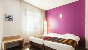 Quarto de hotel com duas camas, um banco, uma janela com cortinas e uma parede roxa.