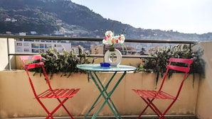 Uma varanda com duas cadeiras vermelhas, uma mesa redonda pequena com um vaso de flores e vista para uma cidade no alto de uma colina.