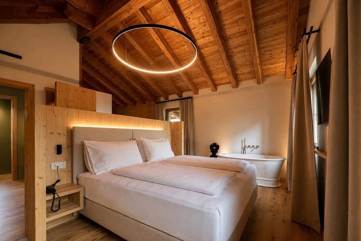Quarto com teto de madeira, cama, mesa de cabeceira, luminária na parede e banheiro com banheira e cortinas.