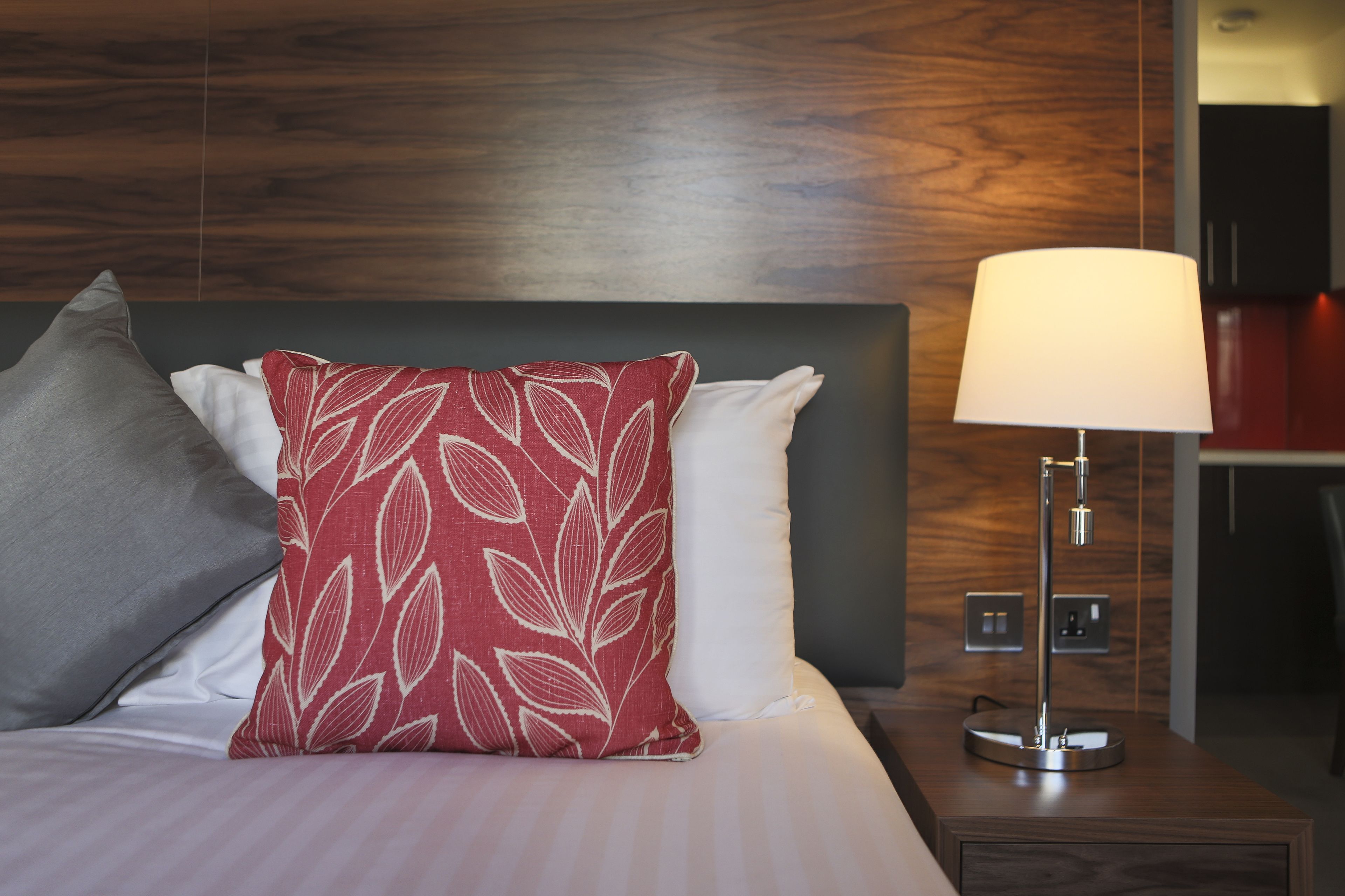 Cama bem arrumada com um travesseiro estampado com folhas vermelhas, um travesseiro branco e um travesseiro cinza. Tem uma mesa de cabeceira com uma luminária e uma tomada.