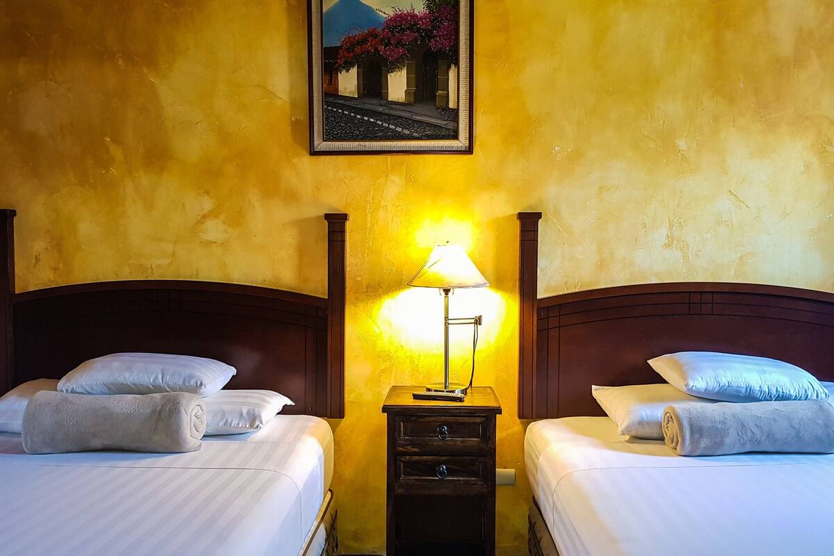 Quarto de hotel com duas camas de solteiro, uma mesinha de cabeceira com abajur e um quadro na parede.
