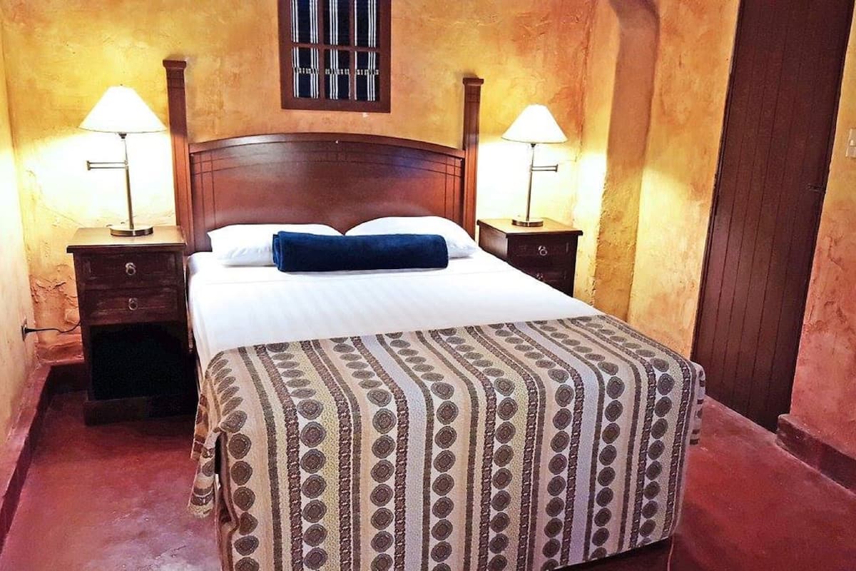 Quarto com cabeceira de madeira, duas mesas de cabeceira com abajures, colcha estampada e janela com grades.