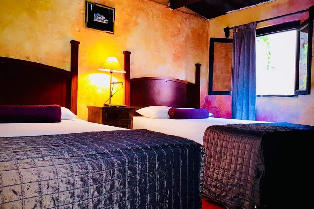 Quarto de hotel com duas camas, uma mesa de cabeceira com abajur e uma janela com cortinas.