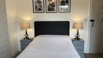 Cama bem arrumada com cabeceira preta, duas mesas de cabeceira com luminárias e três impressões emolduradas de plantas na parede.