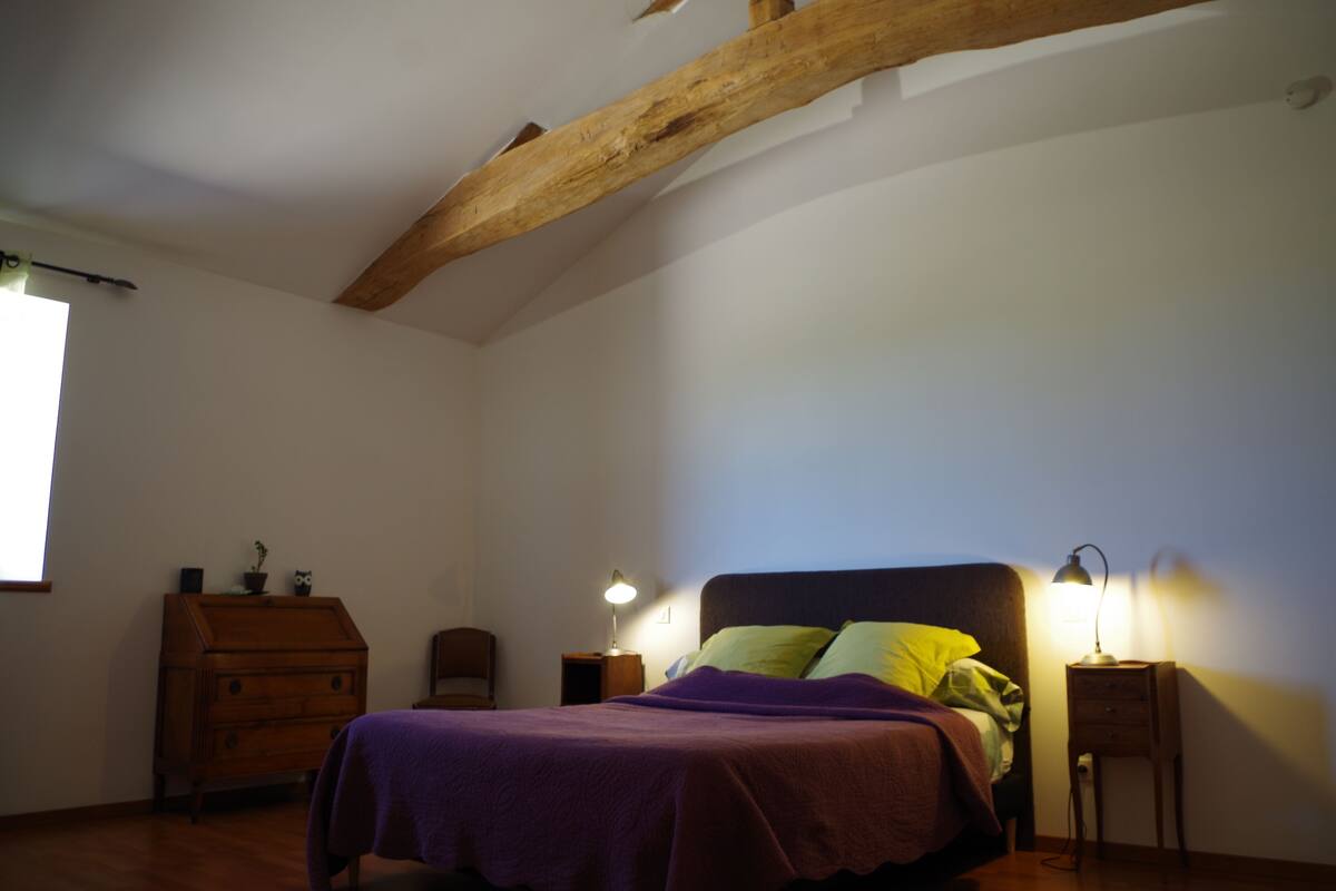 Quarto com uma viga de madeira no teto, uma cama com manta roxa, duas mesas de cabeceira com abajures e um aparador.