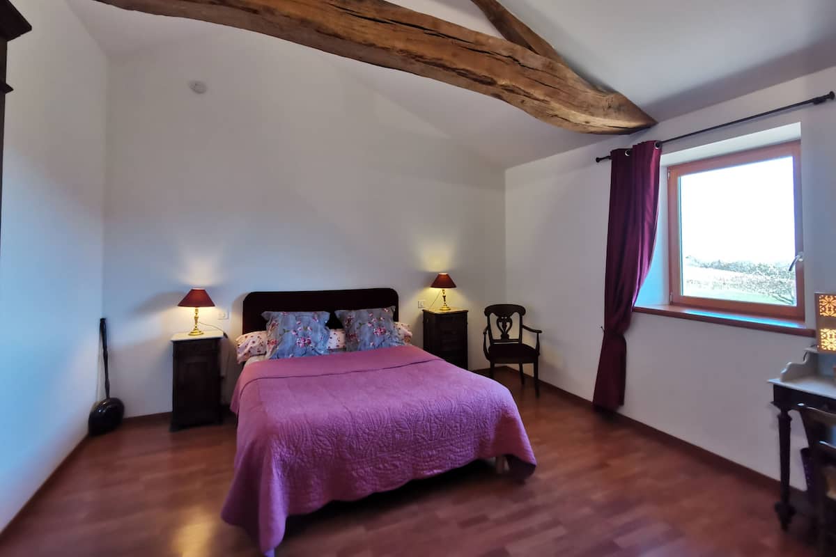 Quarto com teto de madeira, cama com colcha roxa, duas luminárias de apoio, uma cadeira e janela com cortinas.