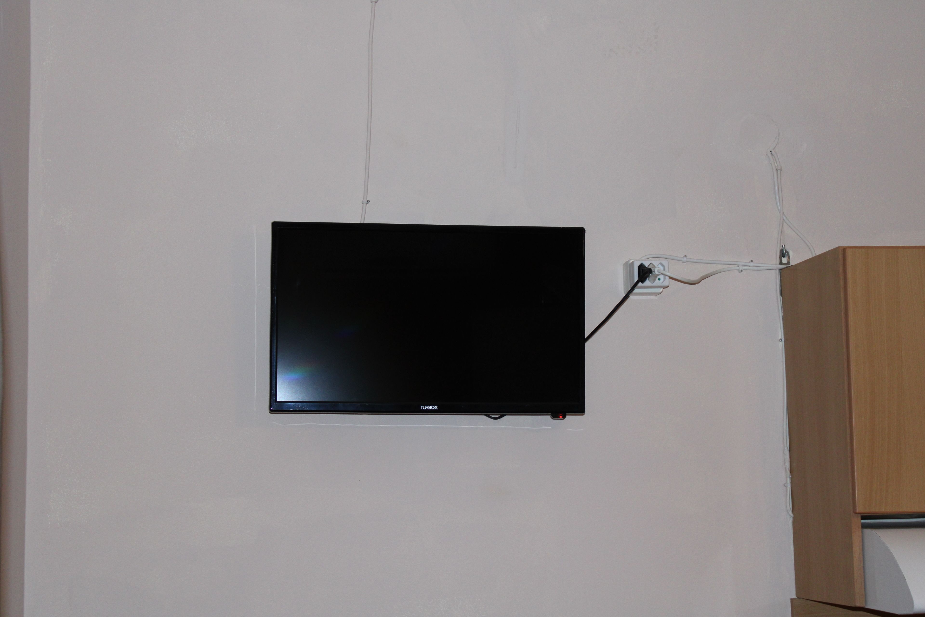 Televisão de tela plana presa em uma parede branca, com um armário de madeira à direita.