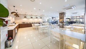 Uma área de refeições moderna com mesas e cadeiras brancas, bancada de cozinha e luminárias pendentes.