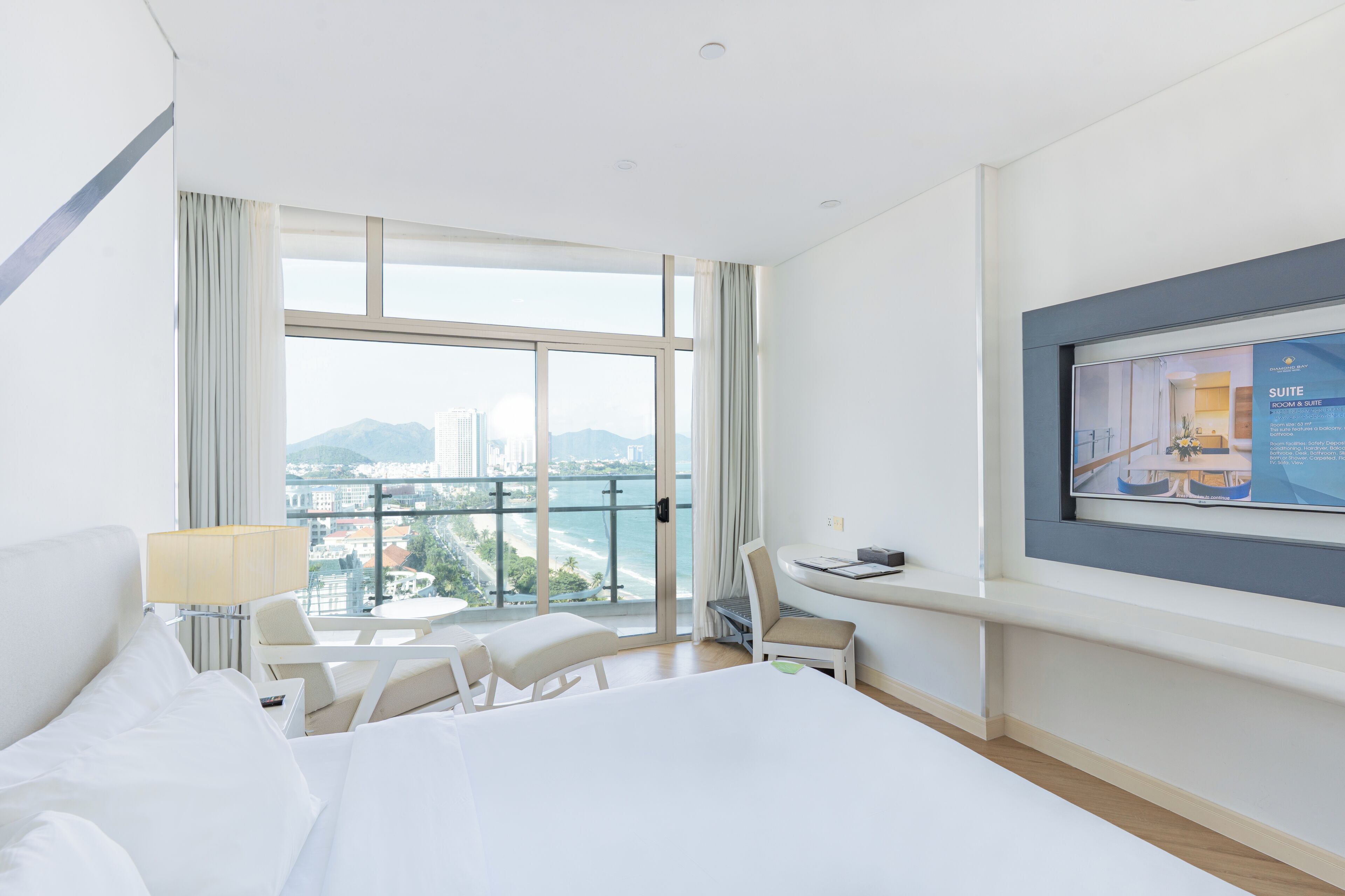 Quarto de hotel com cama, escrivaninha, cadeira, televisão e uma grande janela com vista para a cidade.