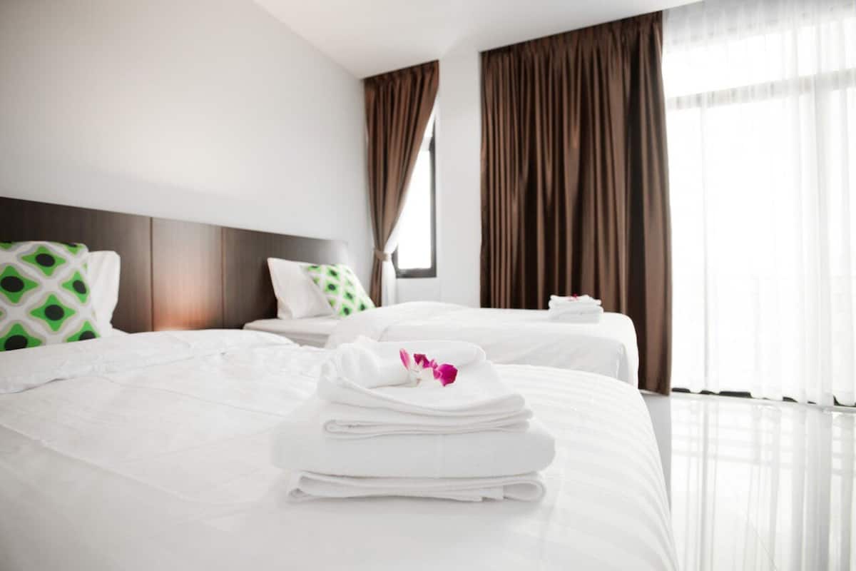Quarto de hotel com duas camas, uma janela grande com cortinas, e uma cama bem arrumada com toalhas e uma flor.