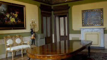 Uma sala formal com uma grande mesa de madeira, cadeiras ricamente decoradas, um candelabro e um quadro clássico.