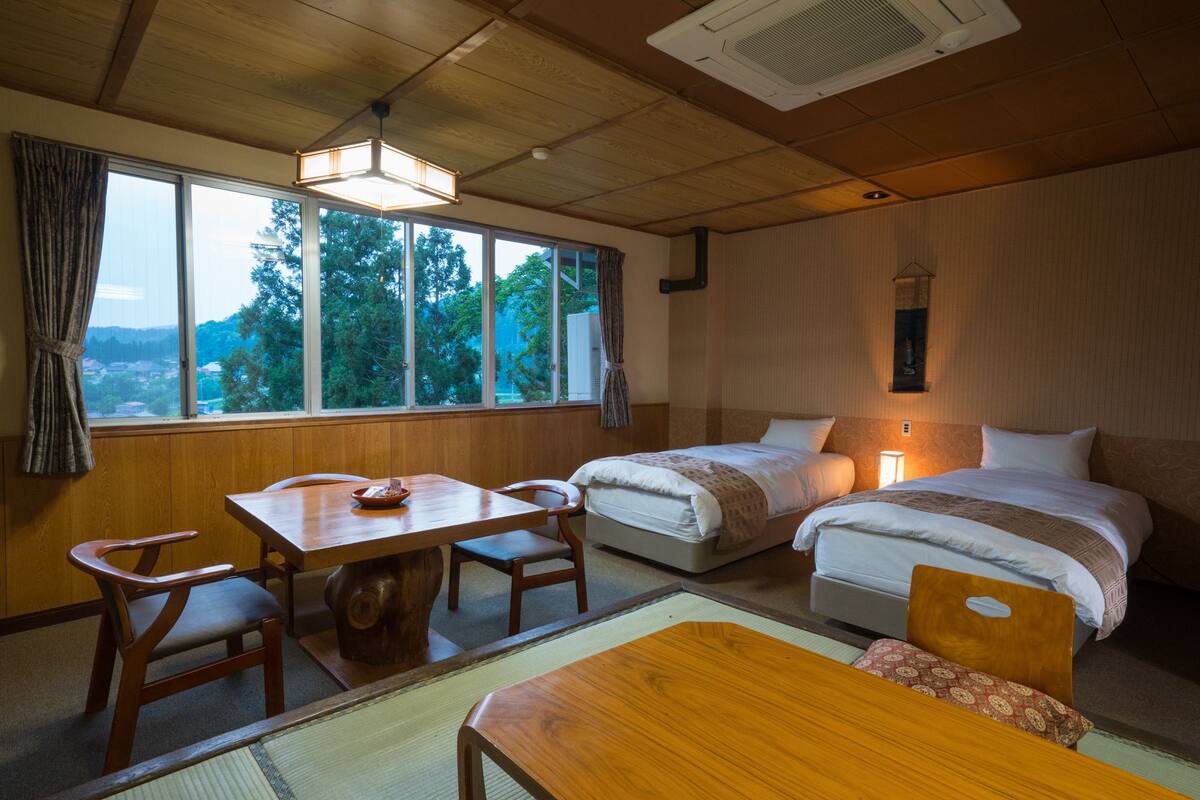 Quarto com teto de madeira, duas camas, uma mesa de jantar com cadeiras e vista para árvores por grandes janelas.