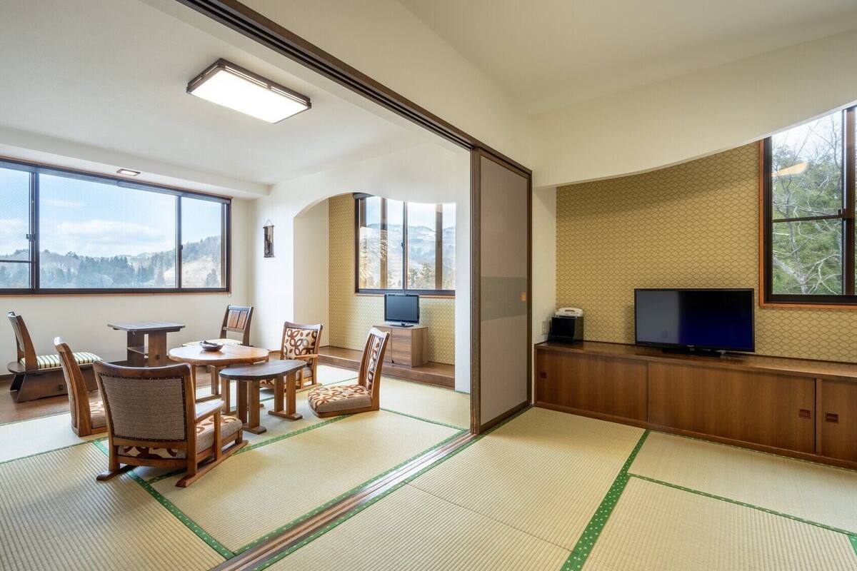Um quarto japonês tradicional com piso de tatame, uma mesa baixa, cadeiras, uma televisão e grandes janelas com vista para o exterior.