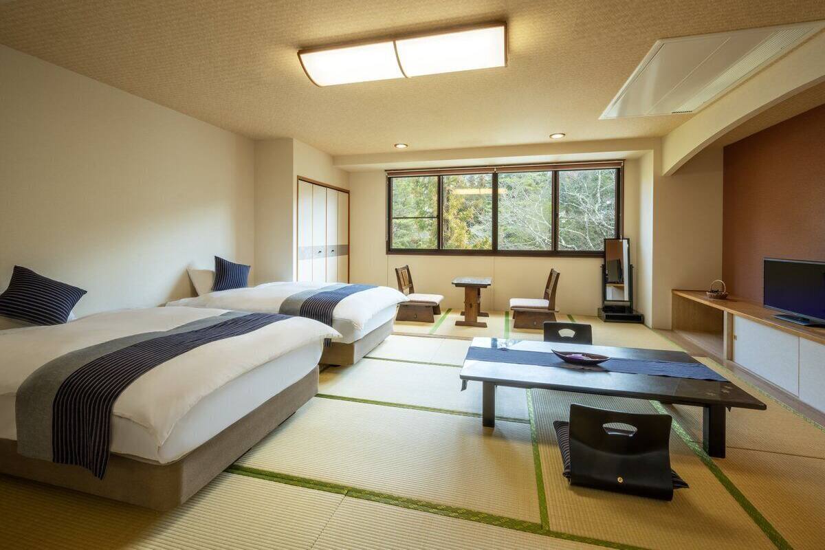 Quarto de hotel com duas camas, uma escrivaninha, uma cadeira, televisão e uma janela grande com vista para árvores.