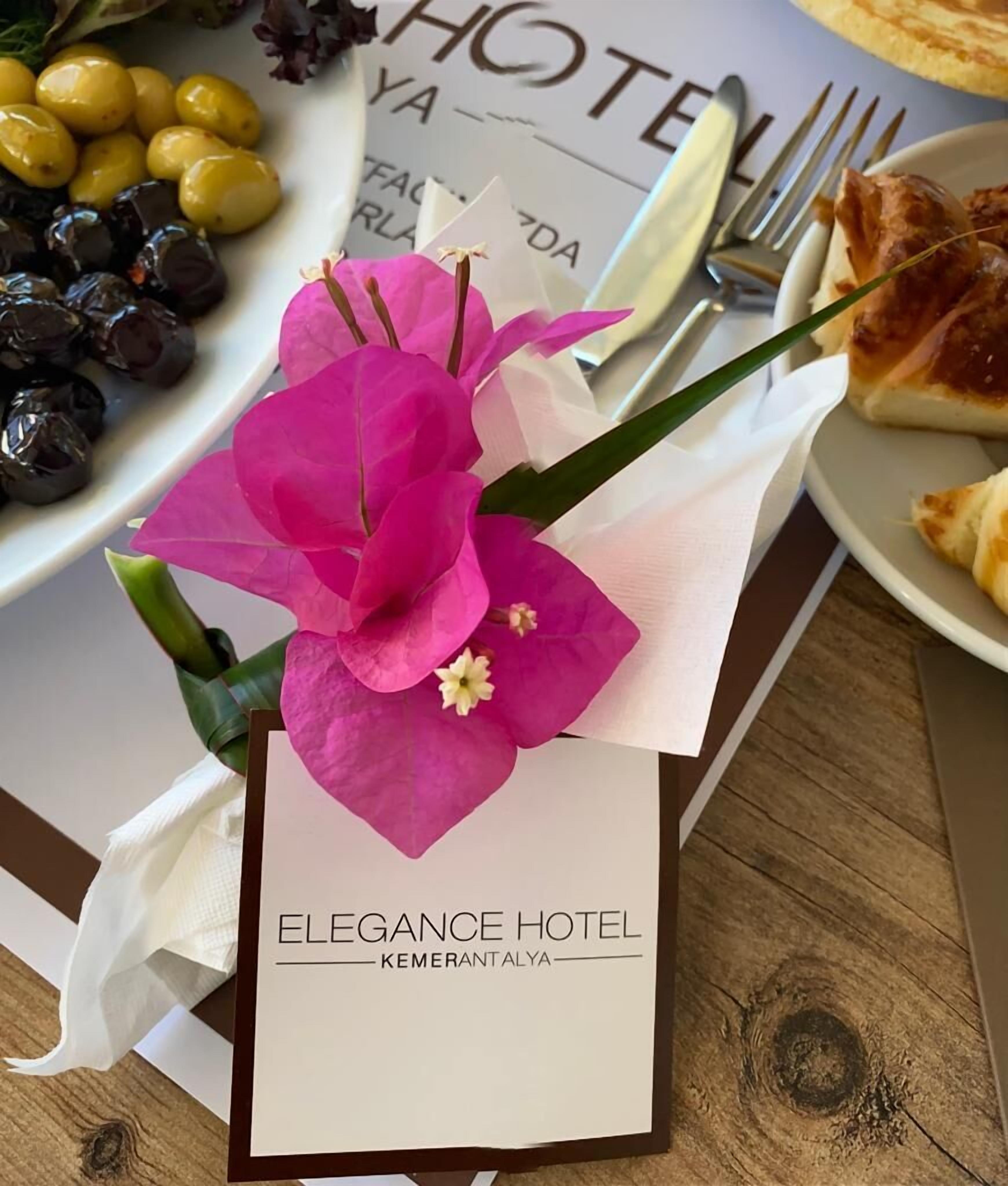 Uma mesa posta com um prato de azeitonas, um arranjo de flores cor-de-rosa e um cartão com o nome do hotel.