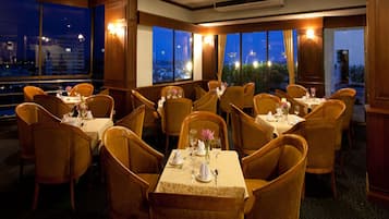 Restaurante com várias mesas e cadeiras, grandes janelas com vista para a cidade e iluminação acolhedora.