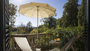 Uma varanda com um guarda-sol bege, uma mesa com frutas e vista para árvores e montanhas.