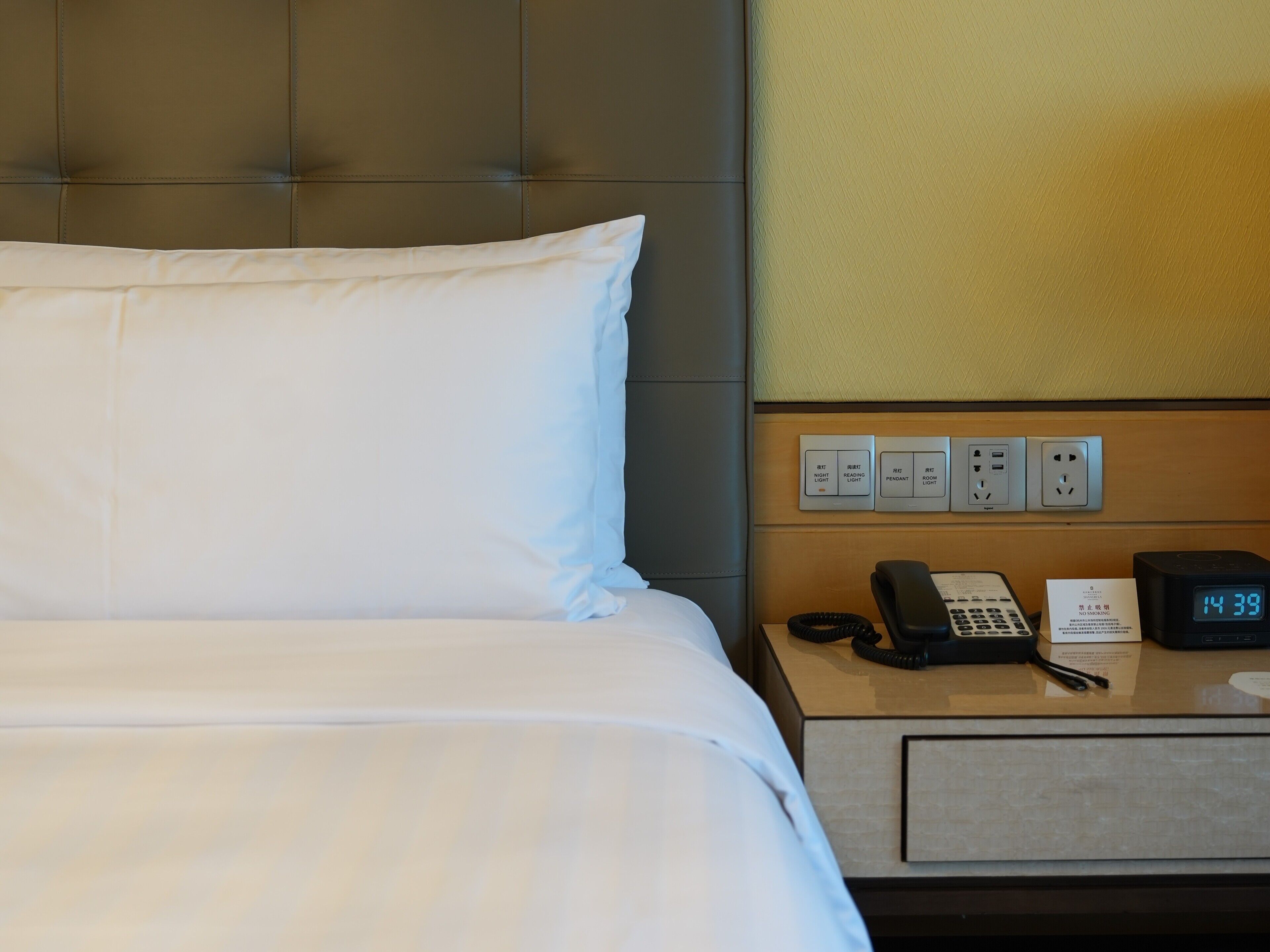 Quarto de hotel com uma cama, mesa de cabeceira com telefone, relógio e tomadas elétricas.