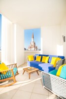 Uma sala de estar iluminada, com um sofá azul, almofadas amarelas, uma cadeira de balanço de madeira e uma cadeira de vime. Um quadro de um castelo está na parede.