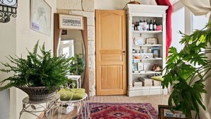 Quarto acolhedor com porta de madeira, prateleira branca com garrafas, tapete estampado e plantas verdes.