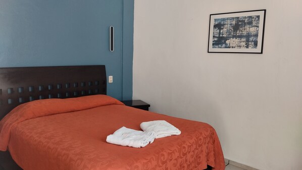 Uma cama com colcha laranja, toalha branca, cabeceira escura, mesa de cabeceira pequena e um quadro abstrato emoldurado na parede.