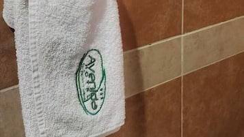 Toalha branca com um logotipo pendurada em um suporte de metal, um cesto com sachês sobre uma superfície branca, e paredes com azulejos marrons.