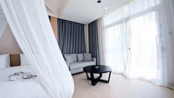 Quarto de hotel moderno com uma cama, um sofá cinza, uma mesa redonda preta e cortinas translúcidas.