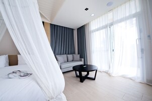 Quarto de hotel moderno com uma cama, um sofá cinza, uma mesa redonda preta e cortinas translúcidas.