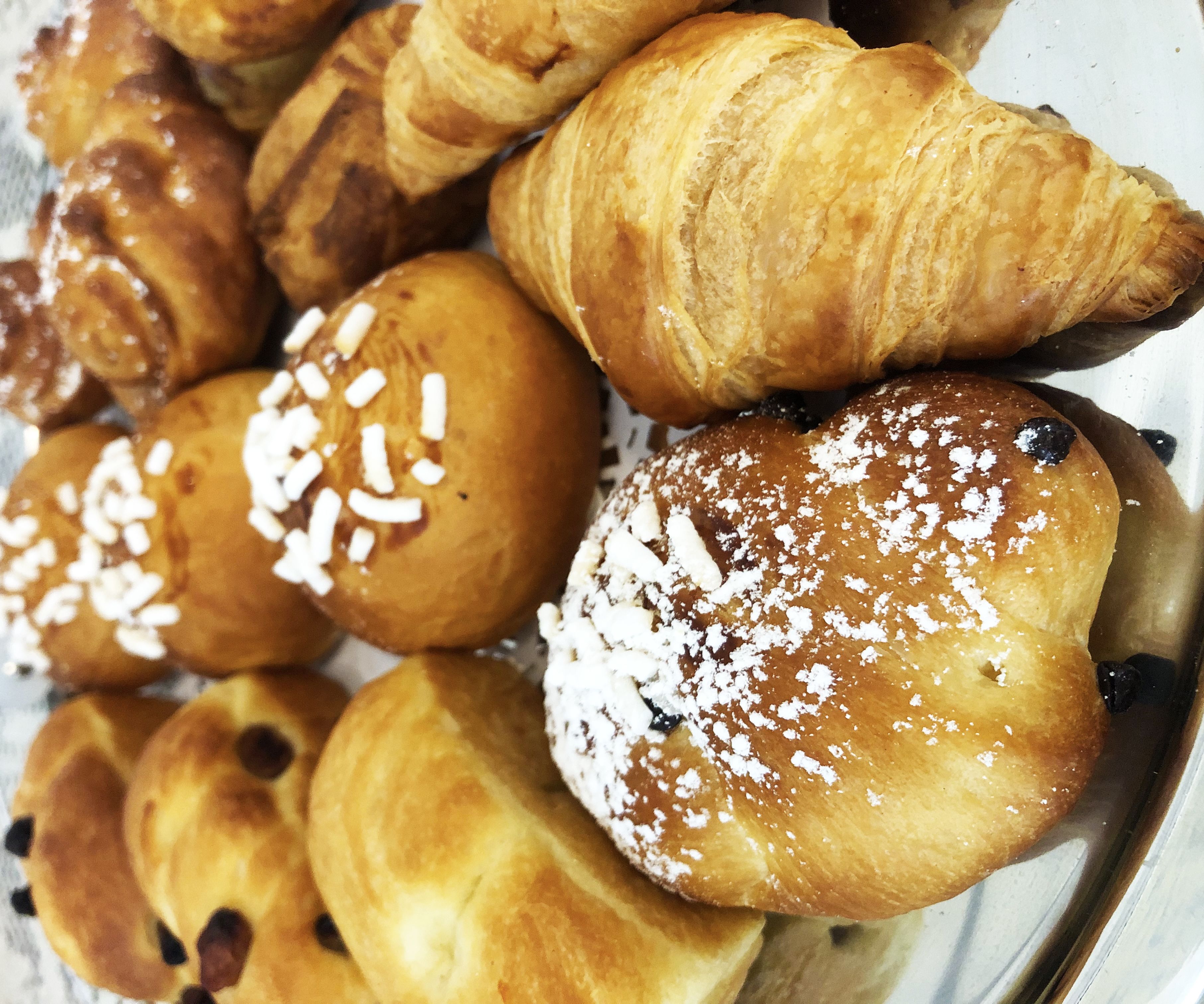 Diversos pastéis, incluindo croissants e pães, alguns cobertos com açúcar em pó e outros com sementes.