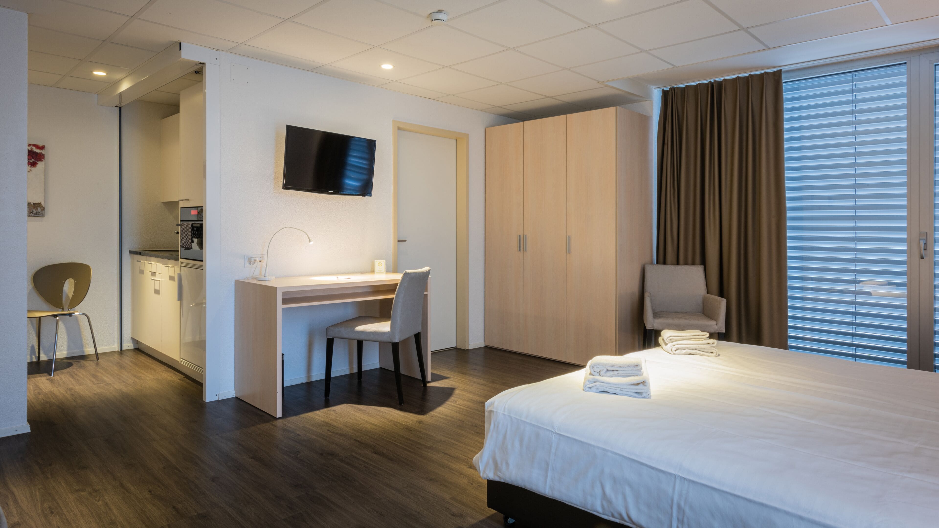 Quarto de hotel com cama, escrivaninha, cadeira, televisão e armário.
