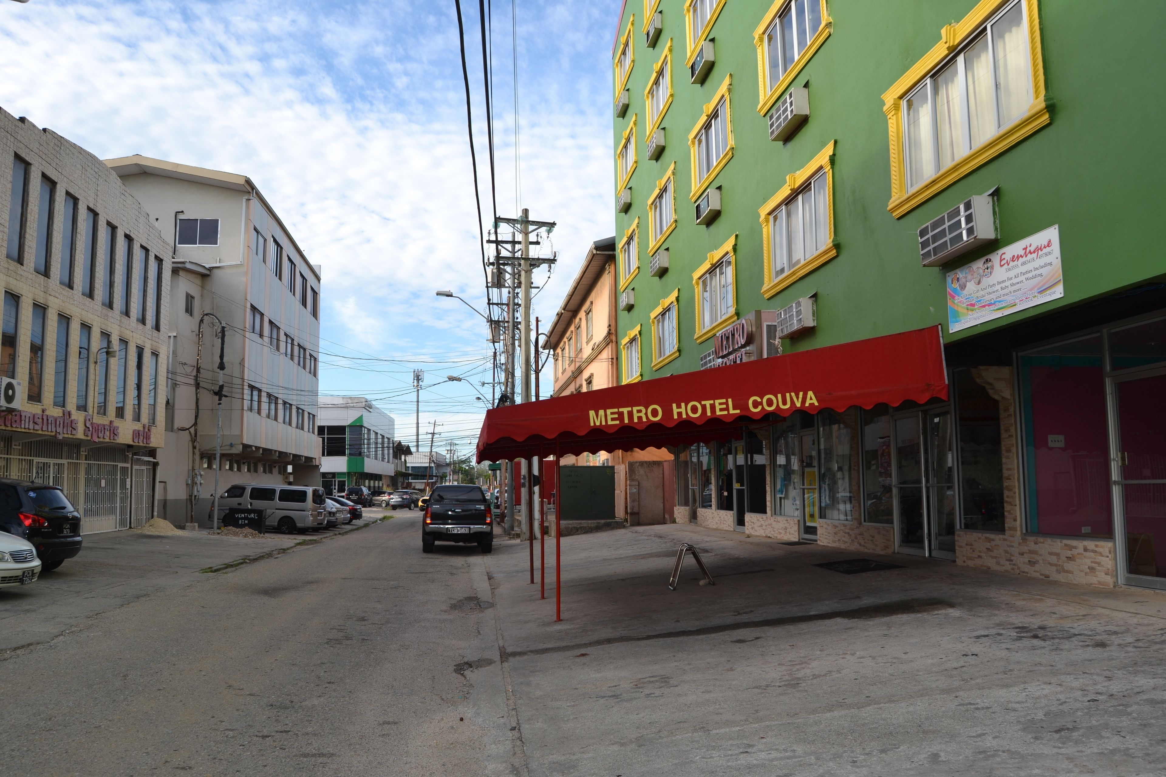 Vista da rua com um hotel chamado "Metro Hotel Cuva", apresentando um toldo vermelho, carros estacionados e edifícios com diversos sinais e janelas.