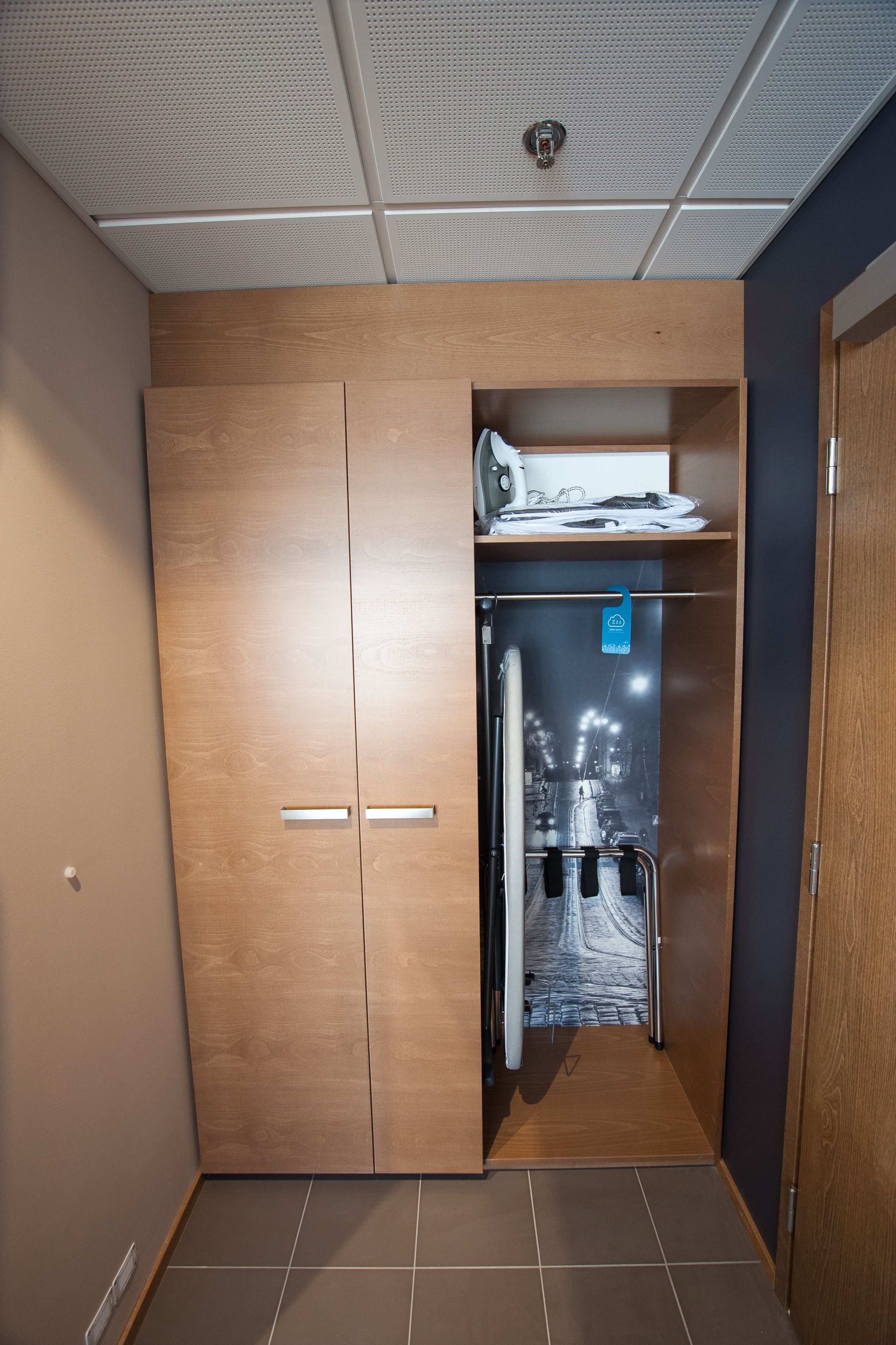 Quarto com guarda-roupa de madeira, uma pequena prateleira com objetos e uma porta com etiqueta azul.