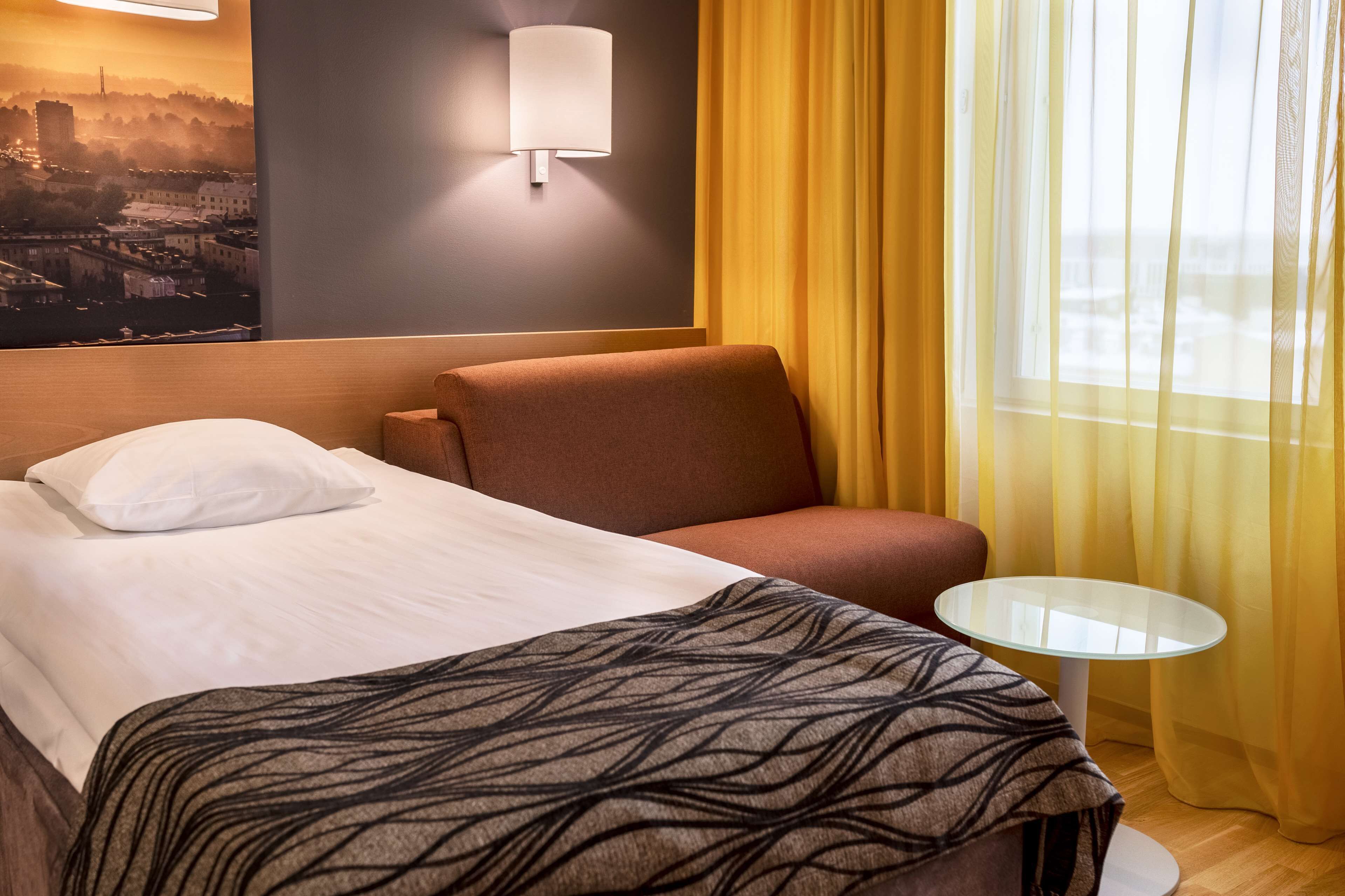 Quarto de hotel com cama, uma cadeira, uma mesinha e uma luminária fixada na parede.