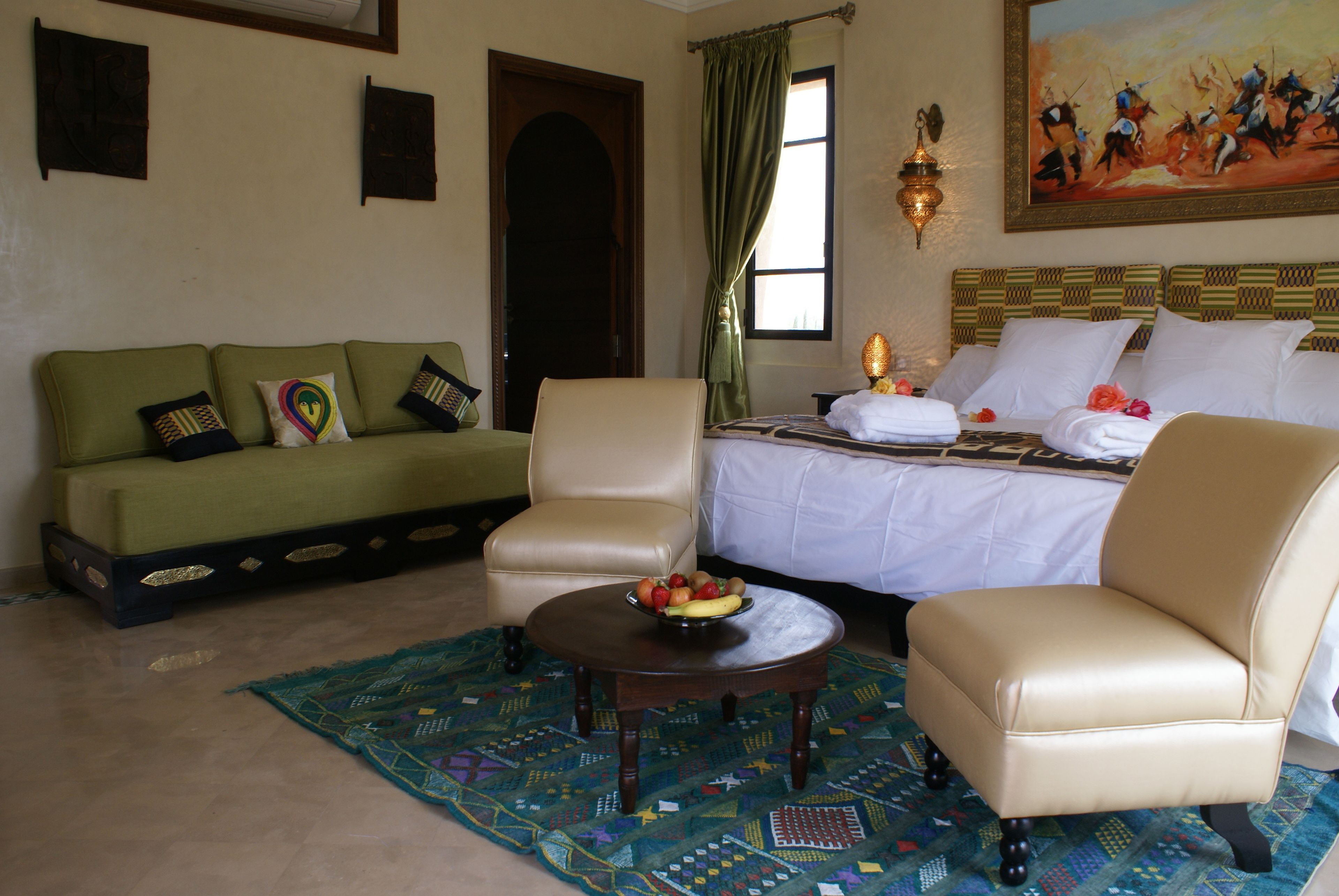 Quarto com sofá verde, cama com lençóis brancos, poltrona bege e mesa redonda com frutas.