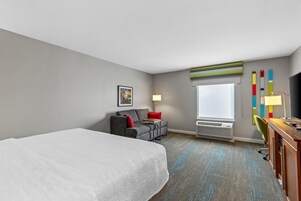 Quarto de hotel com uma cama grande, um sofá cinza com almofadas vermelhas, uma escrivaninha com luminária e uma janela com vista para o exterior.