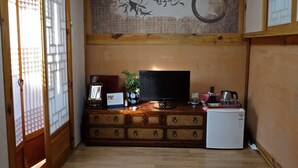 Um quarto japonês tradicional com tatames, um armário de madeira com gavetas, uma televisão, um frigorífico pequeno e uma porta deslizante com painéis translúcidos shoji.