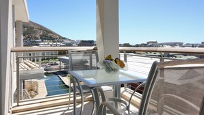 Uma varanda com mesa e cadeiras de vidro, vista para uma piscina e edifícios, e uma montanha ao fundo.