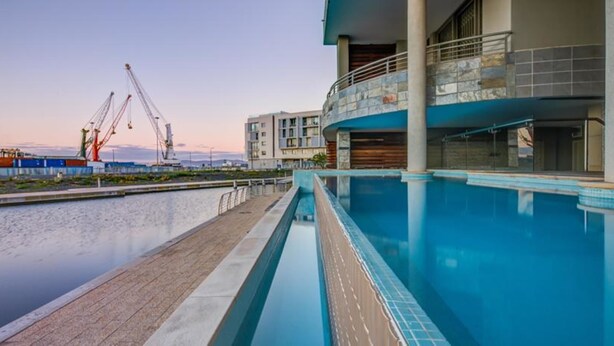 Piscina adjacente a um edifício com varanda, vista para a água e guindastes de construção ao fundo.