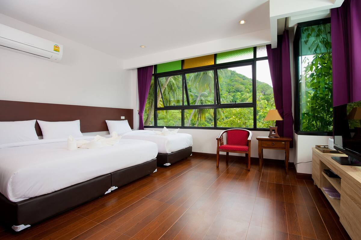Quarto de hotel com duas camas, televisão, uma mesinha e janela com vista para o verde.