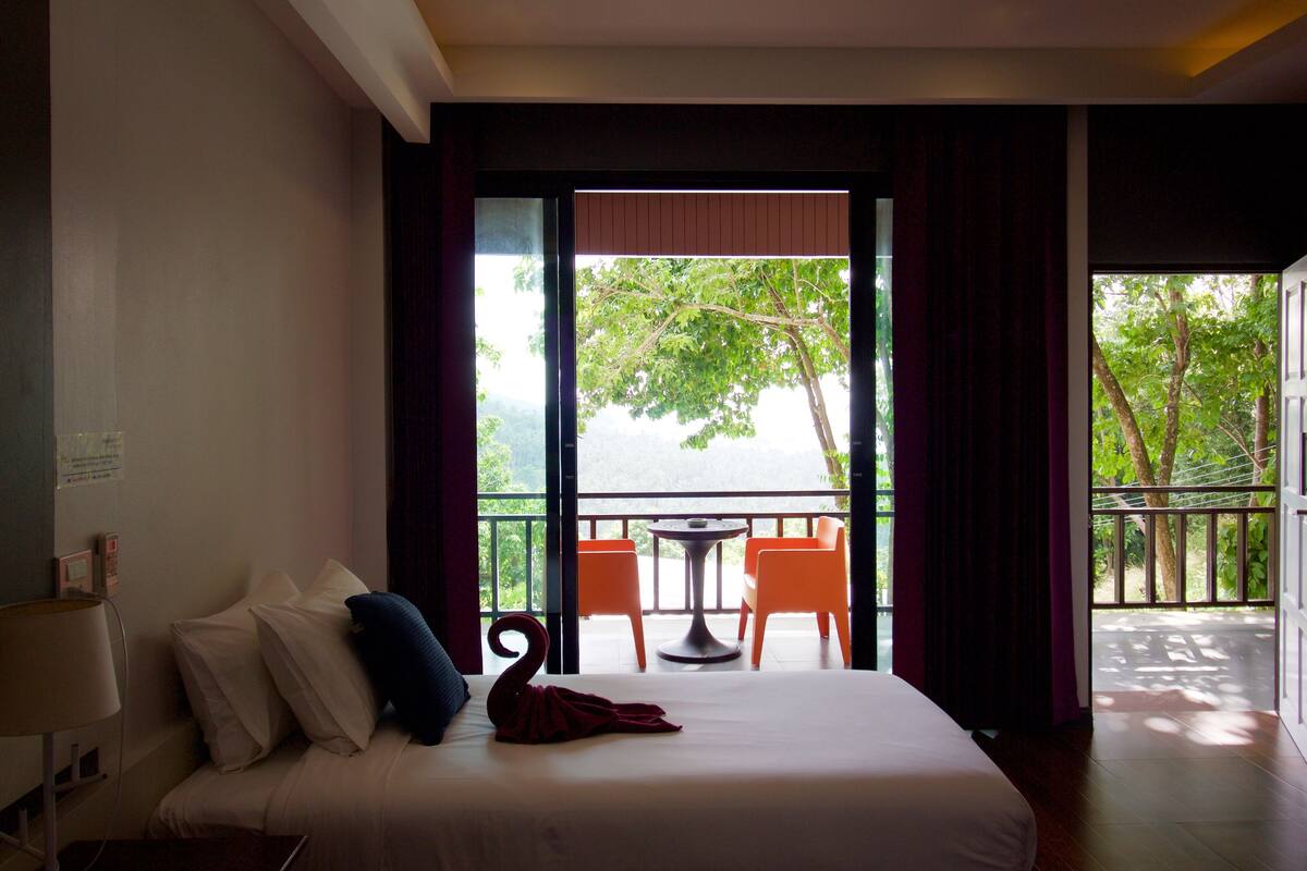 Quarto de hotel com cama, travesseiro em formato de cisne, luminária e varanda com cadeiras e mesa.