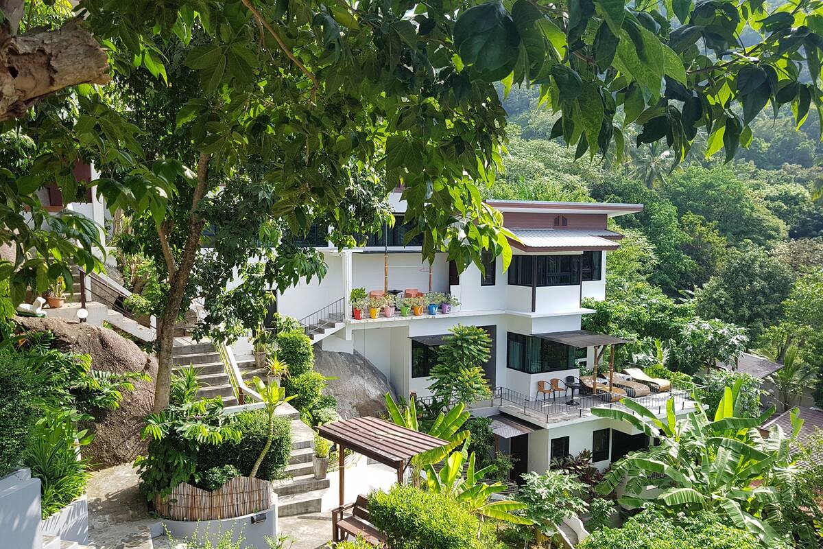 Uma casa moderna de dois andares com uma varanda, cercada por vegetação exuberante e uma escadaria que leva à entrada.
