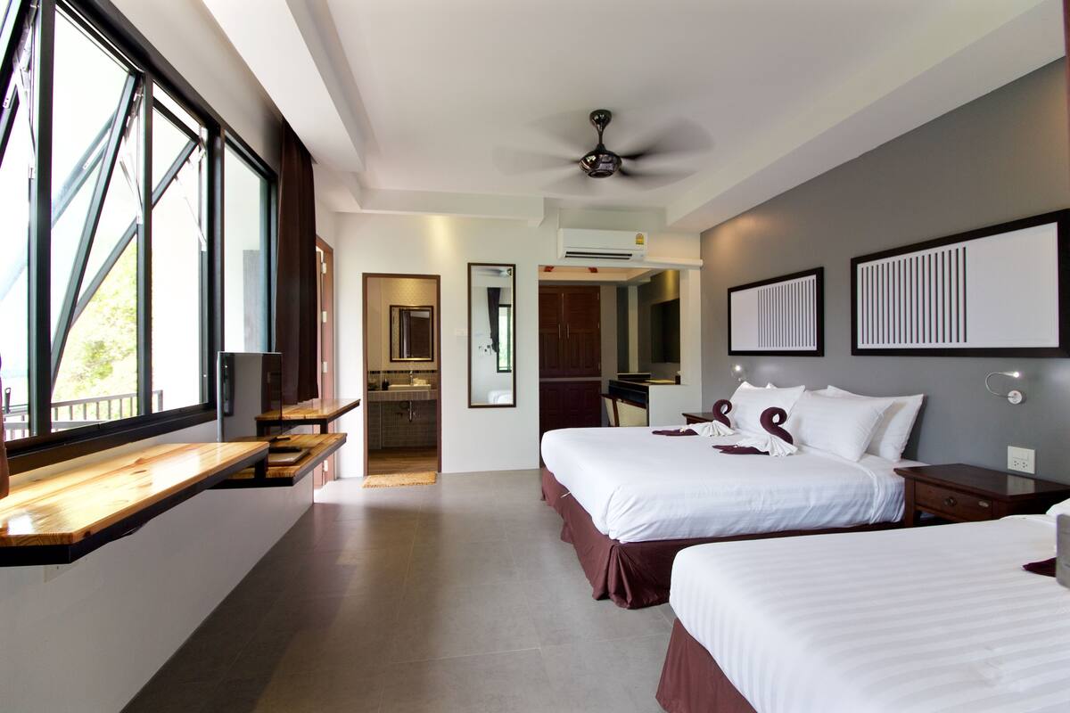 Quarto de hotel com duas camas, uma escrivaninha, um ventilador de teto e uma janela grande.